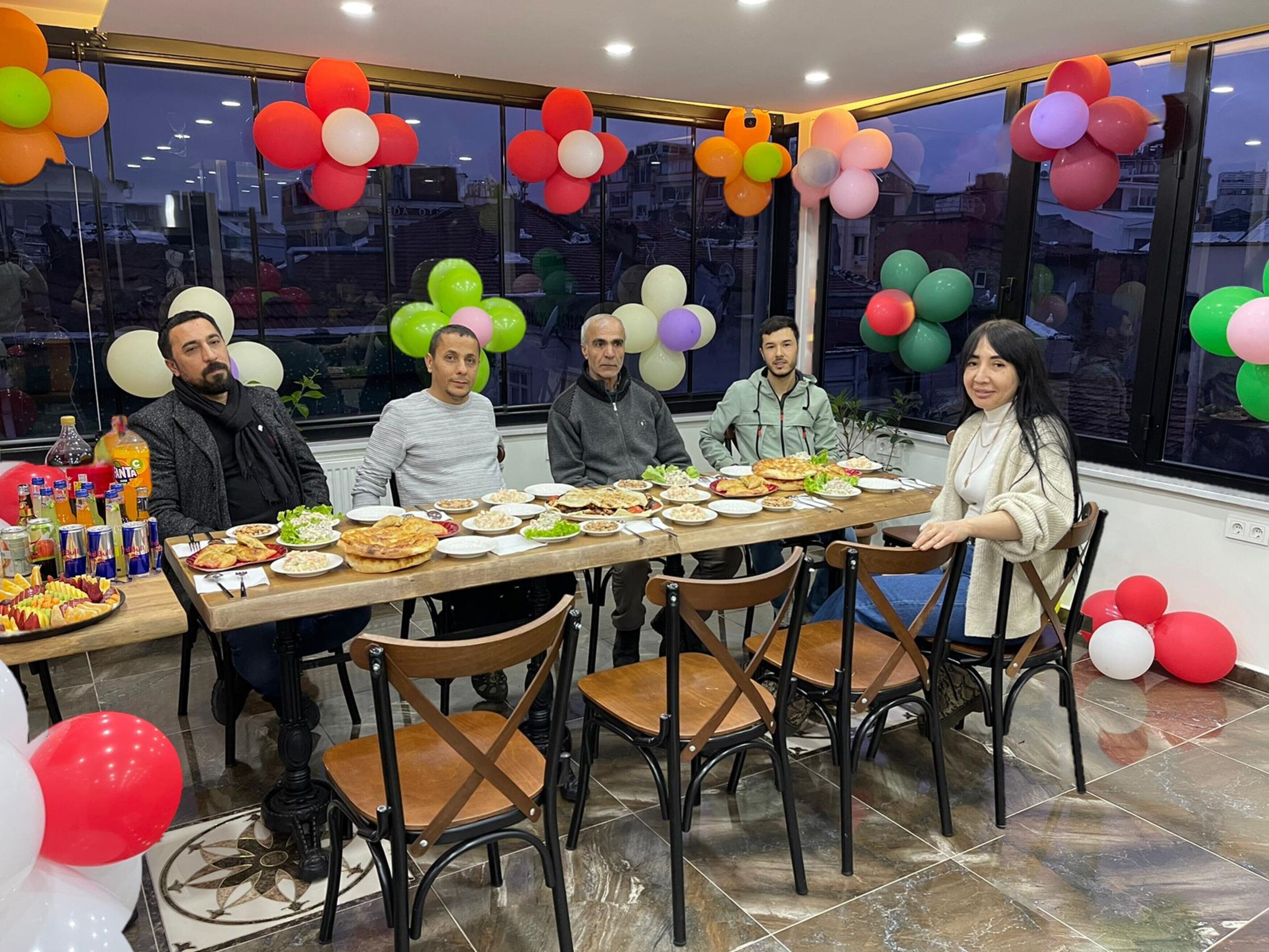 Um grupo de pessoas sentadas à mesa, com vários pratos, balões e vista para a cidade ao fundo.