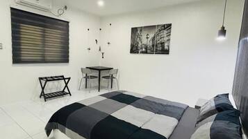 Quarto com uma cama, uma mesinha e cadeiras. Há uma janela com persianas e um ar condicionado fixado na parede.