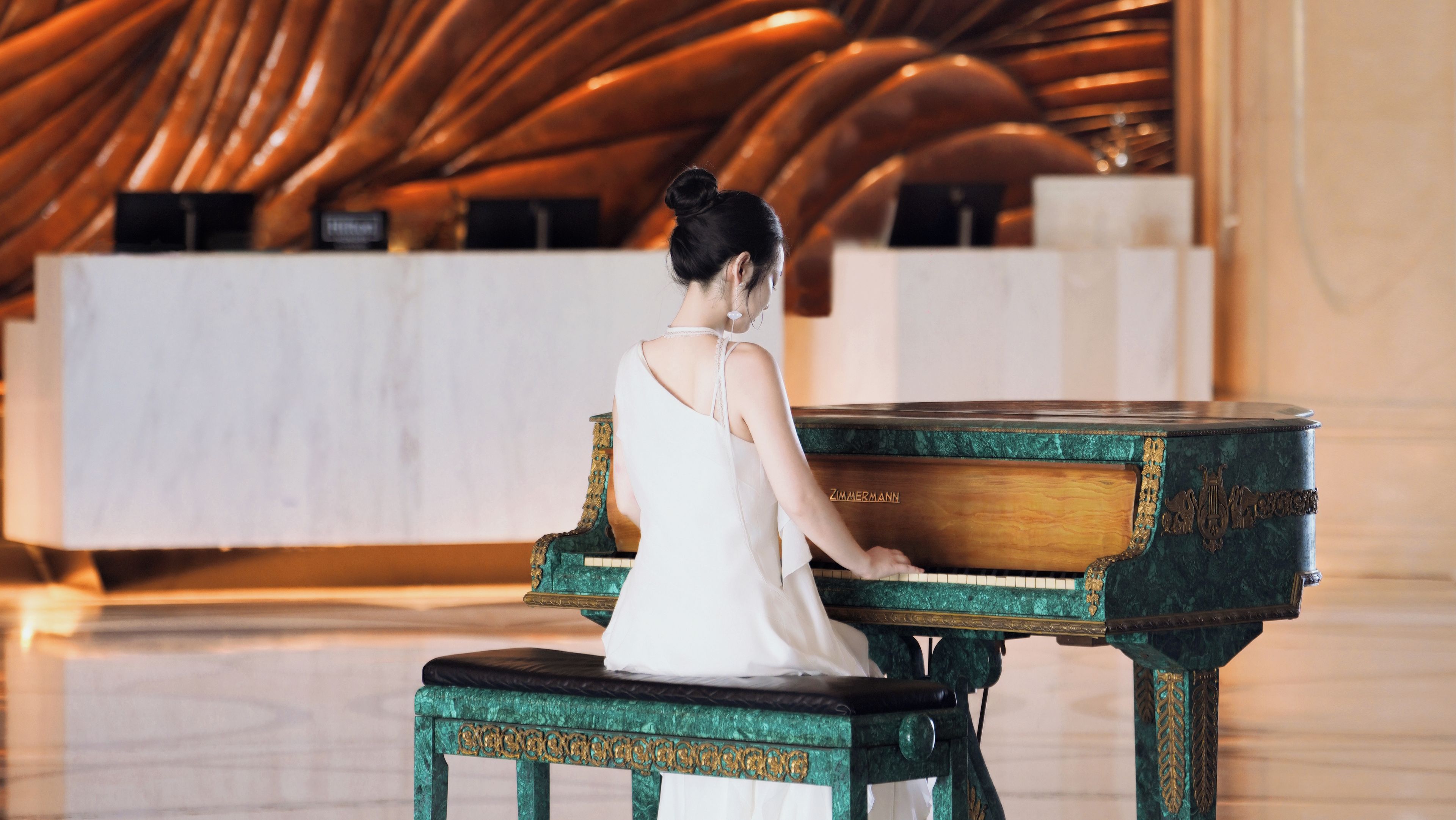 Mulher sentada a um piano de cauda em um amplo saguão, com piso de mármore e design moderno de teto curvo.