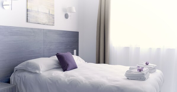 Quarto de hotel moderno com uma cama grande, cabeceira, luminária na parede e uma janela com cortinas.