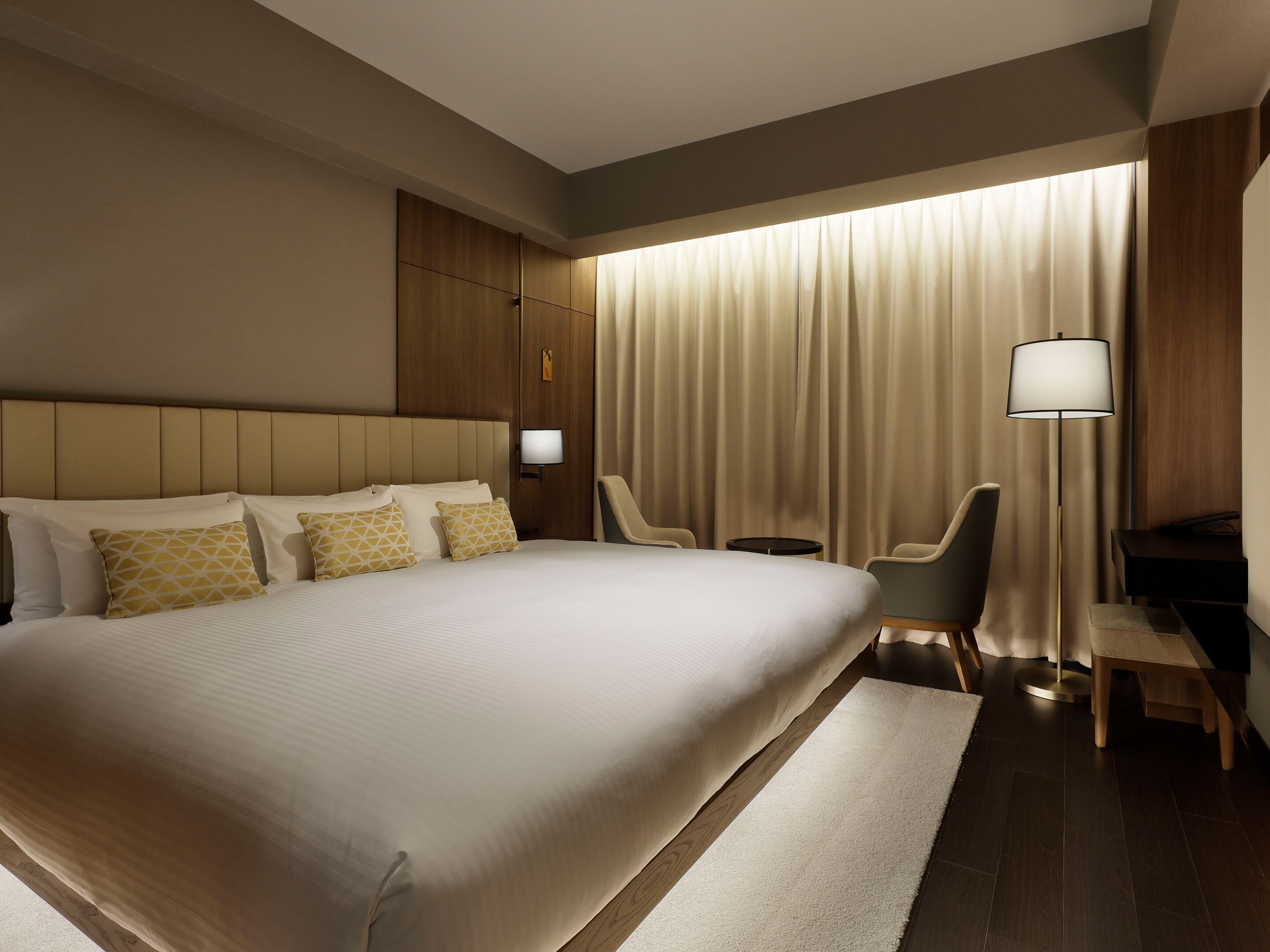 Quarto de hotel com uma cama grande, uma escrivaninha, uma cadeira e um abajur.