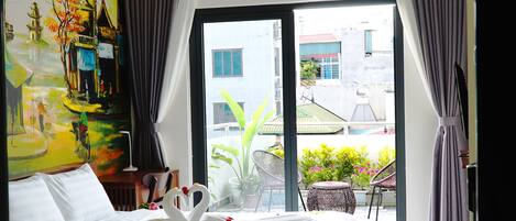 Quarto de hotel com uma cama grande, um quadro na parede, varanda com plantas e vista para prédios.