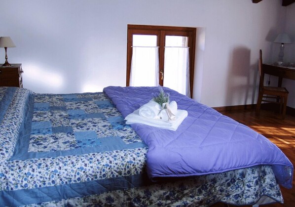 Uma cama bem arrumada com colcha florida azul e edredom roxo, uma mesinha de cabeceira de madeira com abajur, e uma janela com cortinas.