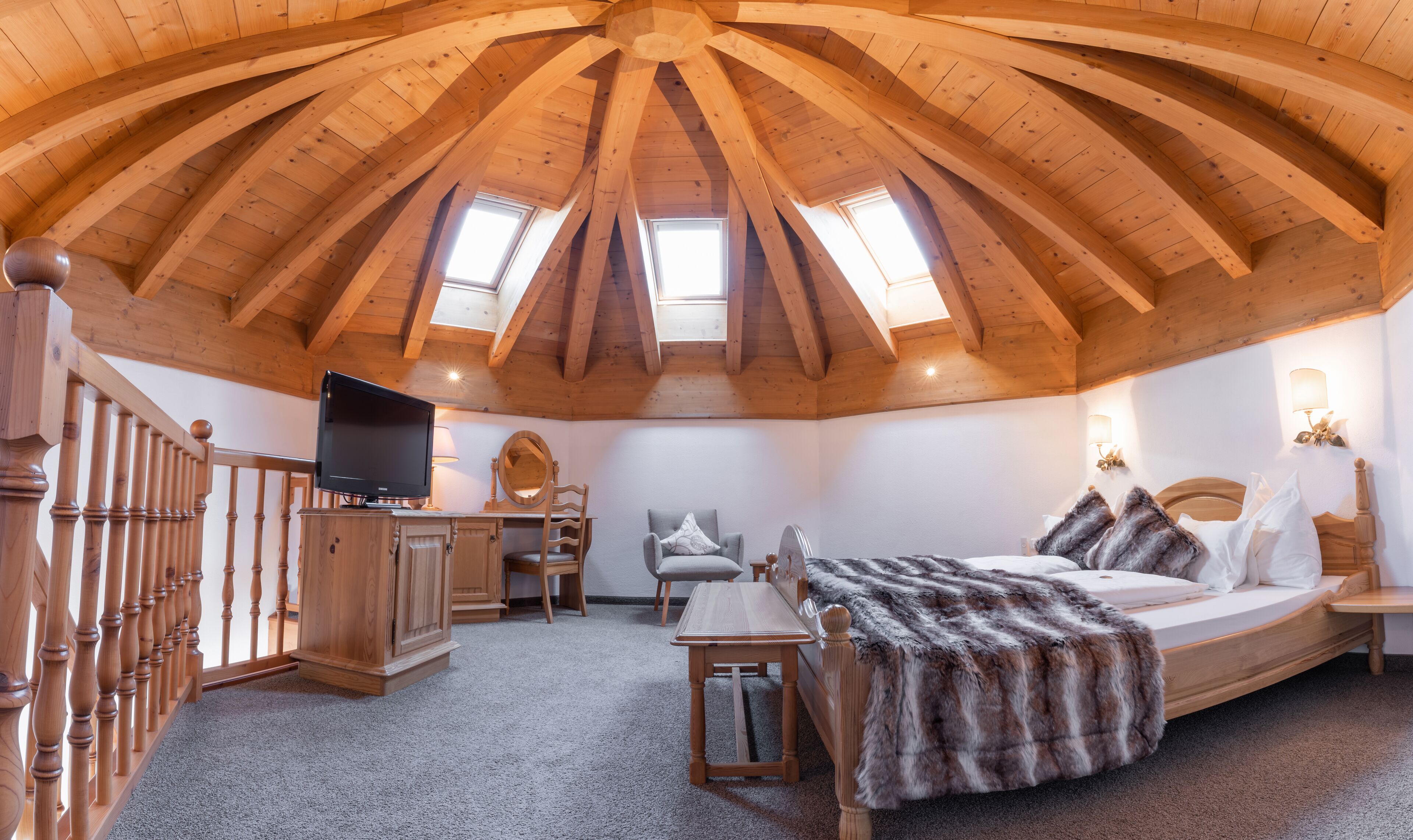 Um quarto com pé direito alto, teto de madeira, cama com manta de pele, escrivaninha com televisão e uma cadeira.