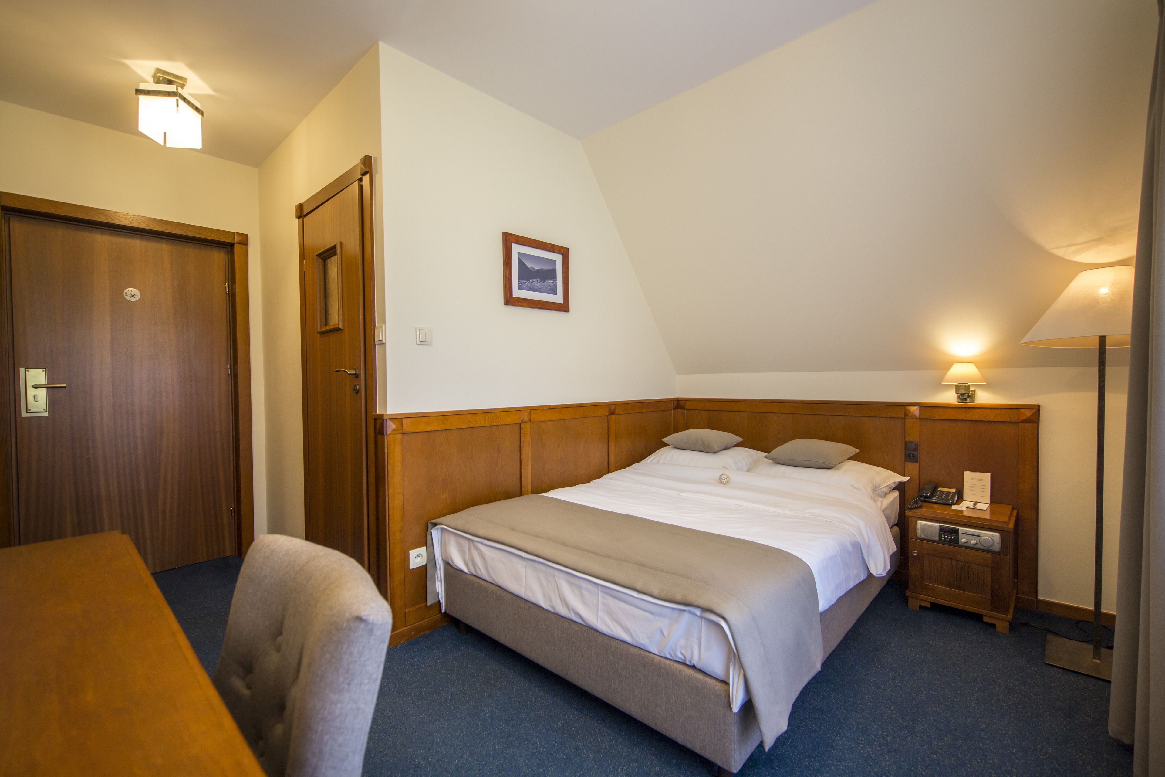 Quarto de hotel com uma cama de solteiro, uma escrivaninha, uma cadeira, mesa de cabeceira com abajur e um quadro na parede.