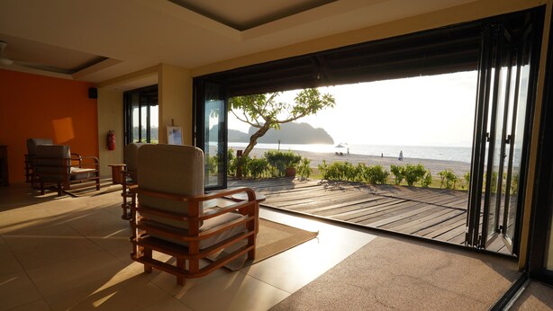 Quarto com cadeira de madeira, vista para a praia e uma grande porta de correr de vidro.