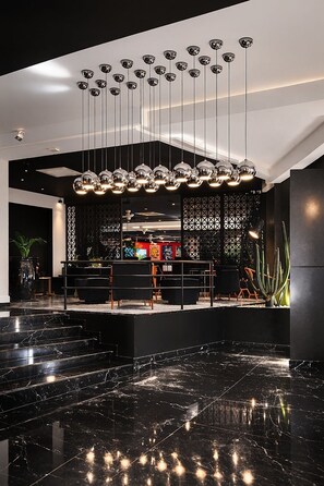 Um hall moderno com piso de mármore preto, balcão de recepção e uma luminária de teto diferenciada.