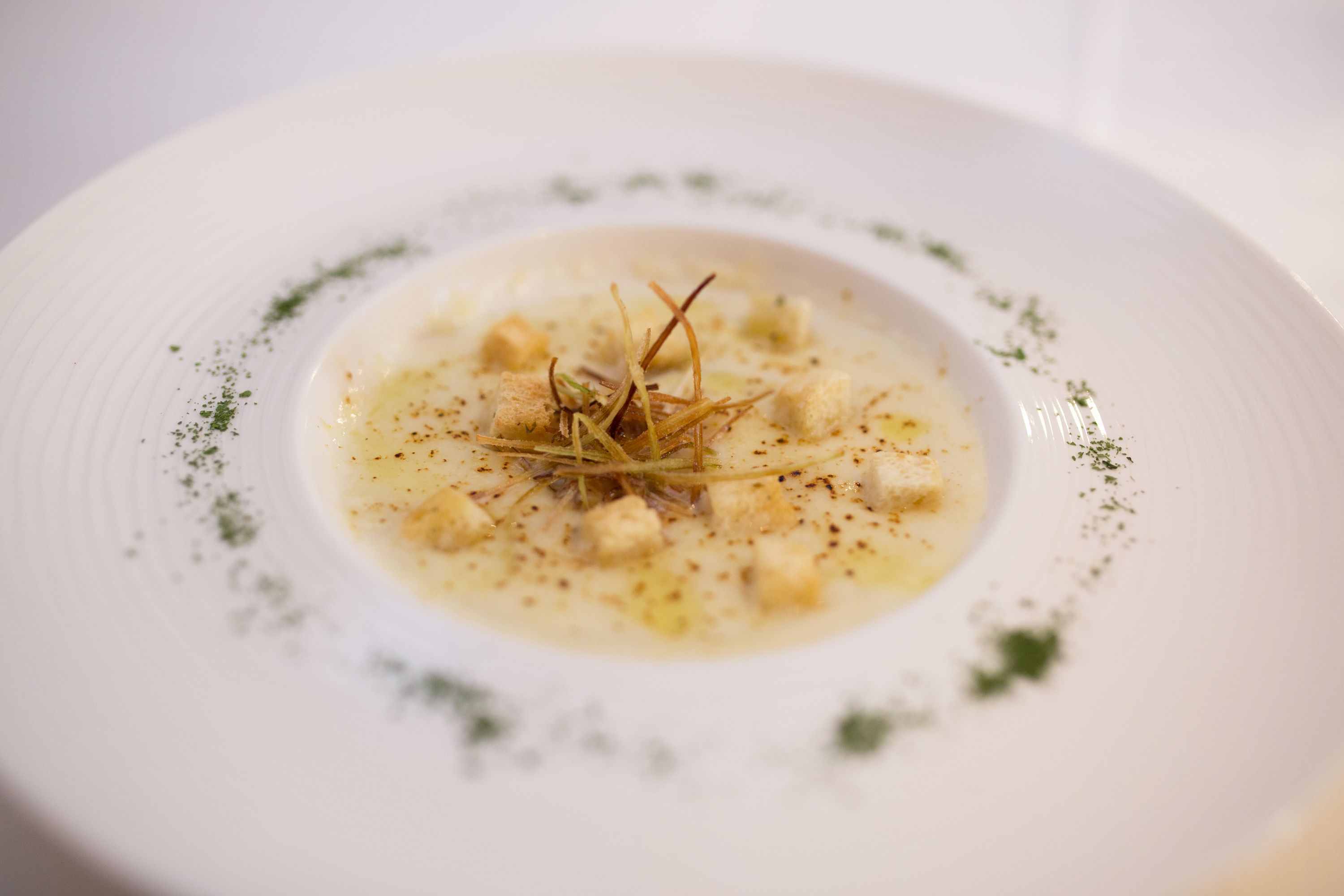 Um prato branco com sopa cremosa, decorado com ervas e um elemento decorativo no centro.