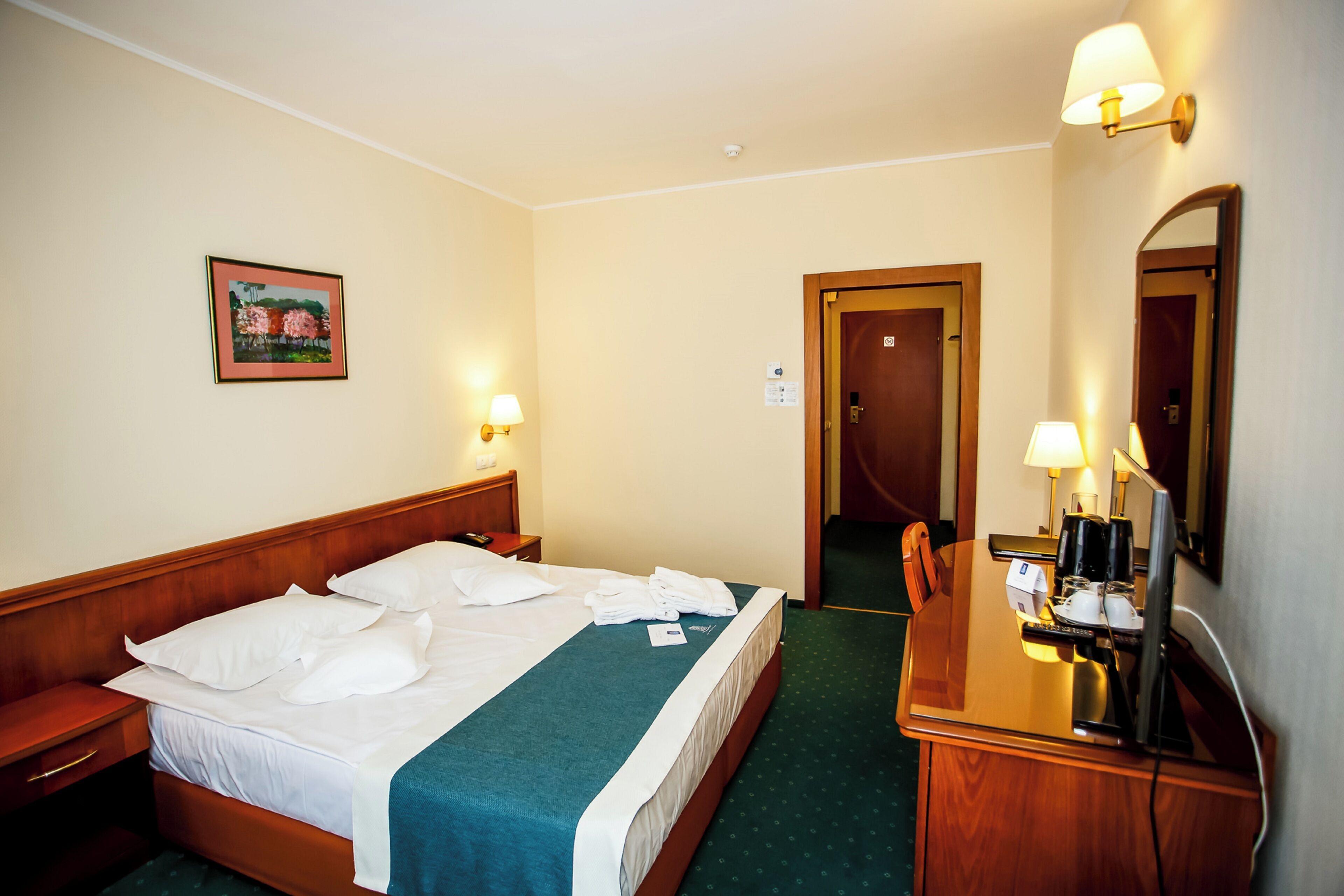 Quarto de hotel com cama, mesas de cabeceira, escrivaninha com cadeira, espelho e um quadro na parede.