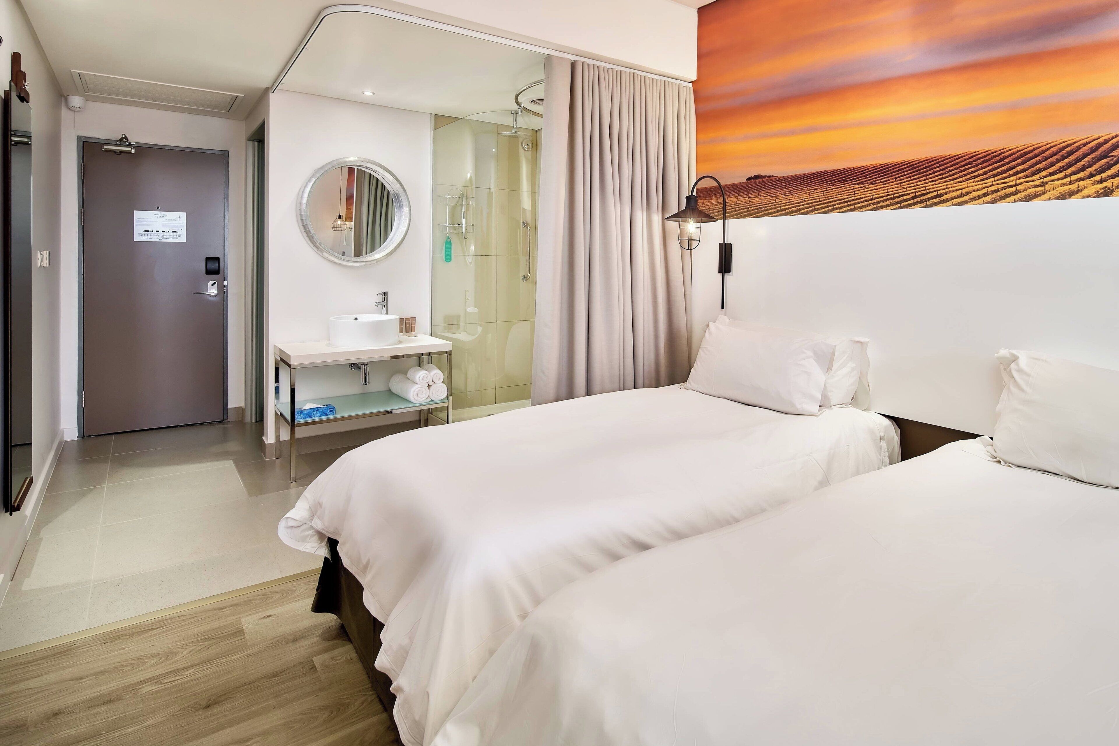 Quarto de hotel com duas camas, banheiro com chuveiro, pia e uma janela grande com vista para o pôr do sol.