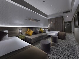 Quarto de hotel moderno com uma cama grande, um sofá, uma mesa redonda pequena e um espaço de estar com um tapete estampado.