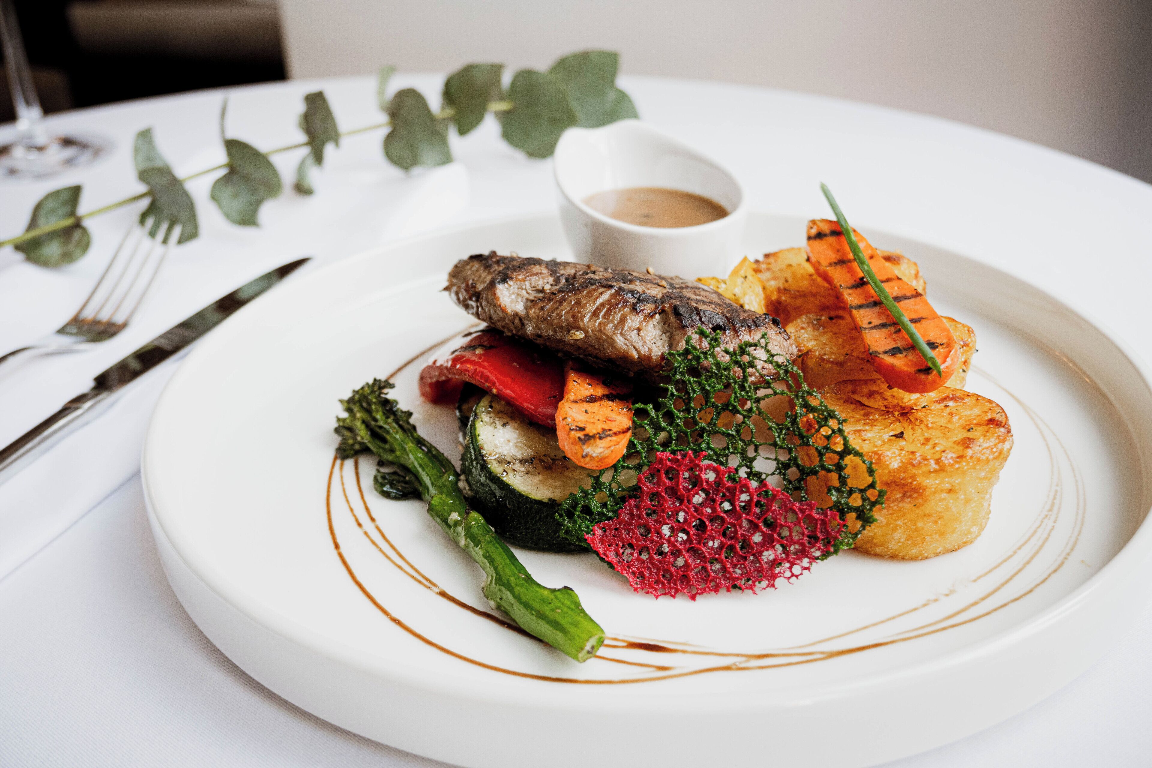 Um prato branco com peixe grelhado, legumes e um detalhe decorativo vermelho, acompanhado de uma pequena tigela de molho.