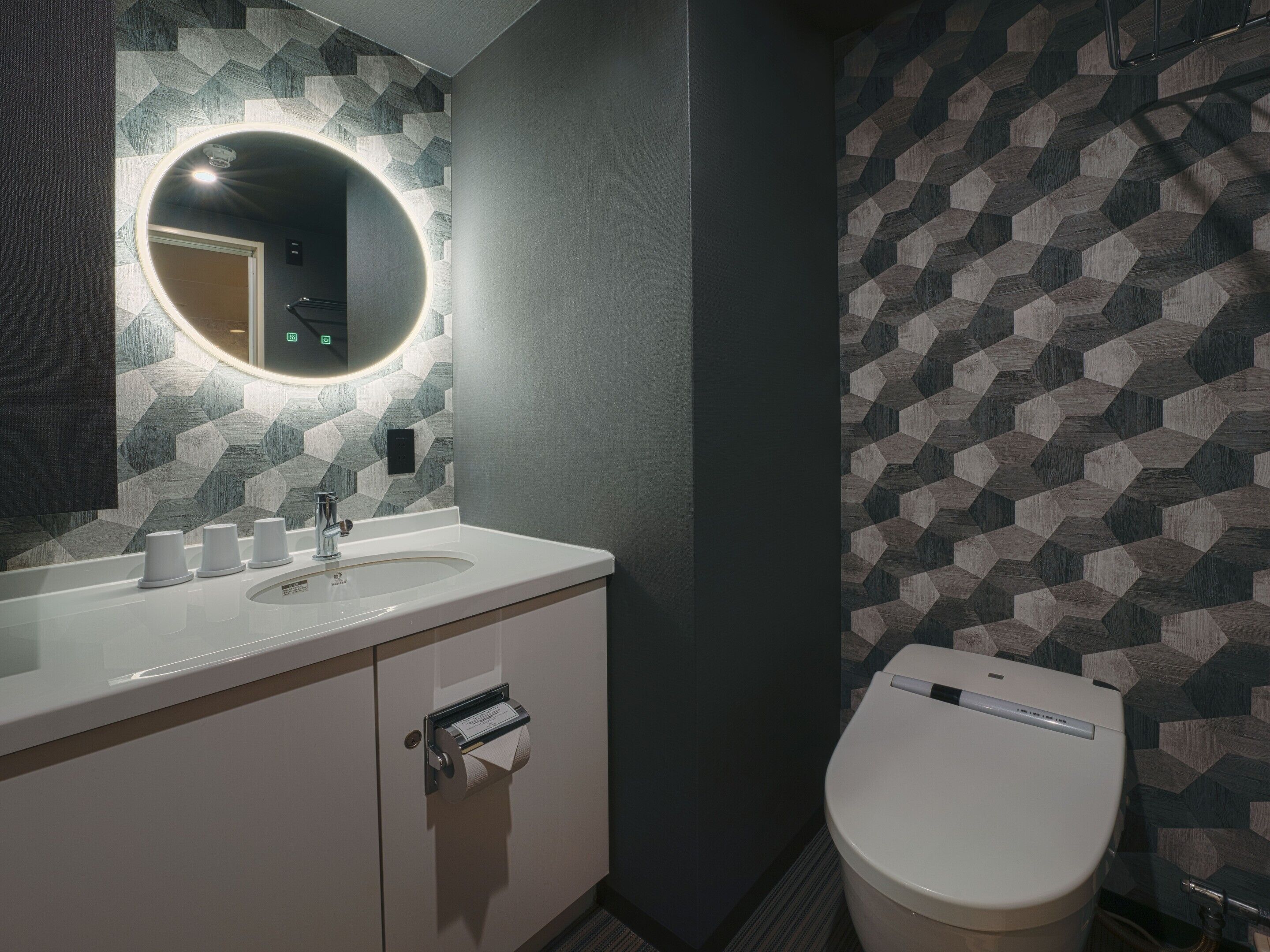 Um banheiro moderno com parede estampada em padrão geométrico, um espelho redondo com iluminação embutida, uma pia branca e um vaso sanitário.
