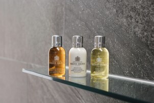 Três frascos de produtos Molton Brown numa prateleira de vidro em frente a uma parede texturizada.