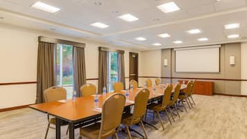 Uma sala de conferência com uma mesa comprida, cadeiras, uma tela de projetor e janelas com cortinas.