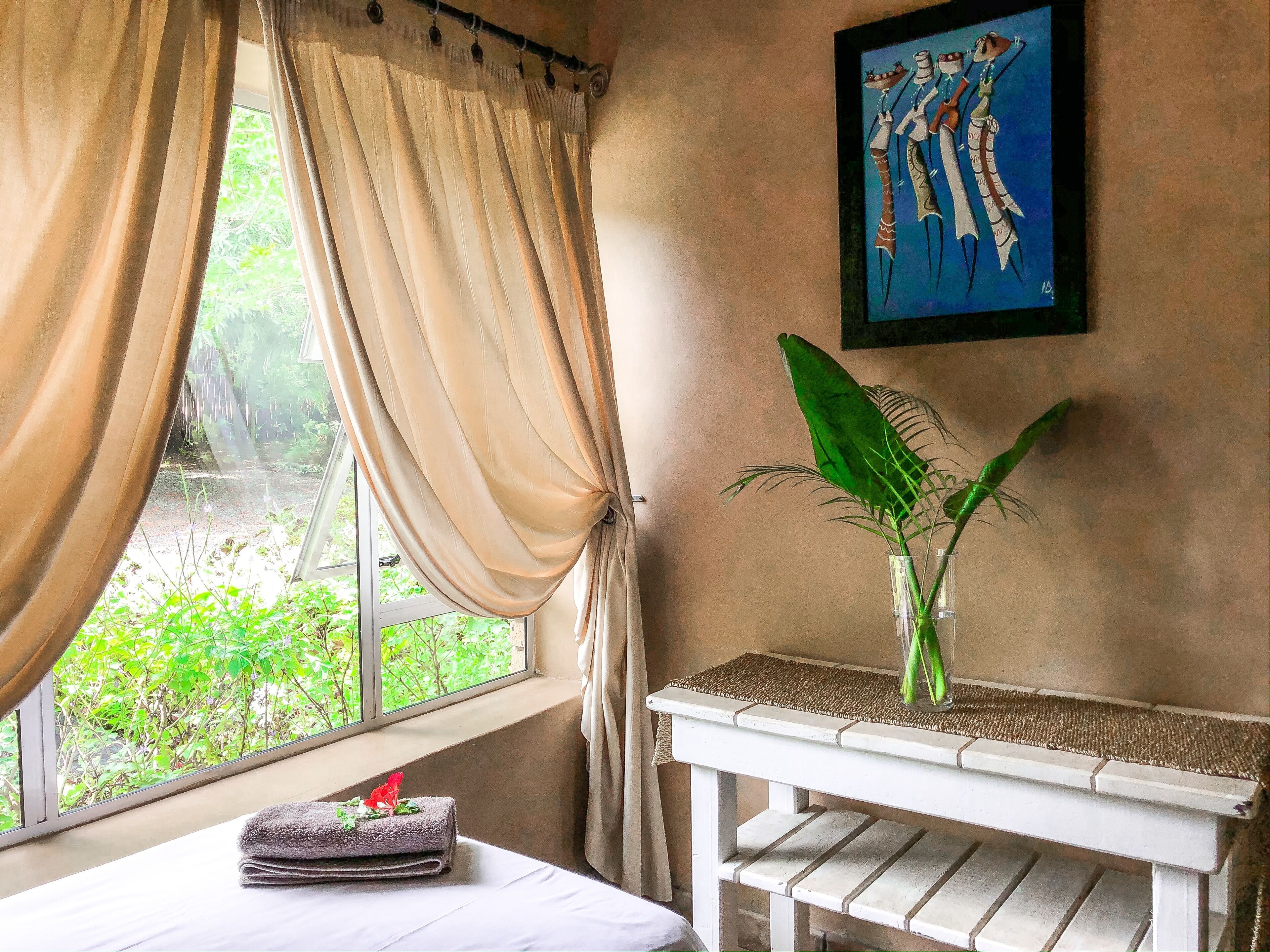 Quarto com cama, janela com cortinas, pintura na parede e planta sobre uma mesa.