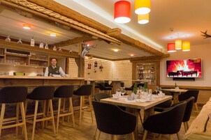 Interior de restaurante com vigas de madeira, área de bar e televisão exibindo uma cena de fogueira.