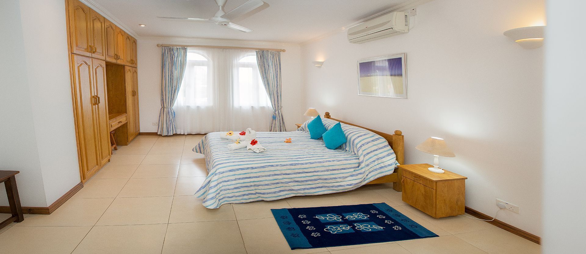 Quarto com cama, mesa de cabeceira, guarda-roupa de madeira, ventoinha no teto e janela com cortinas.