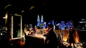 Um quarto com iluminação suave e vista para o horizonte da cidade, incluindo as Torres Petronas, uma mesa com um vaso de flores e uma pessoa próxima à janela.