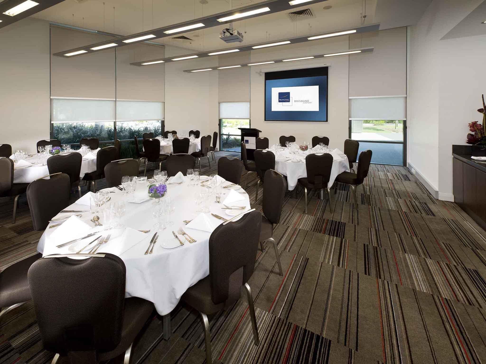 Uma sala de conferência com mesas redondas, cadeiras e uma tela de projeção exibindo o logotipo do Novotel Brisbane.
