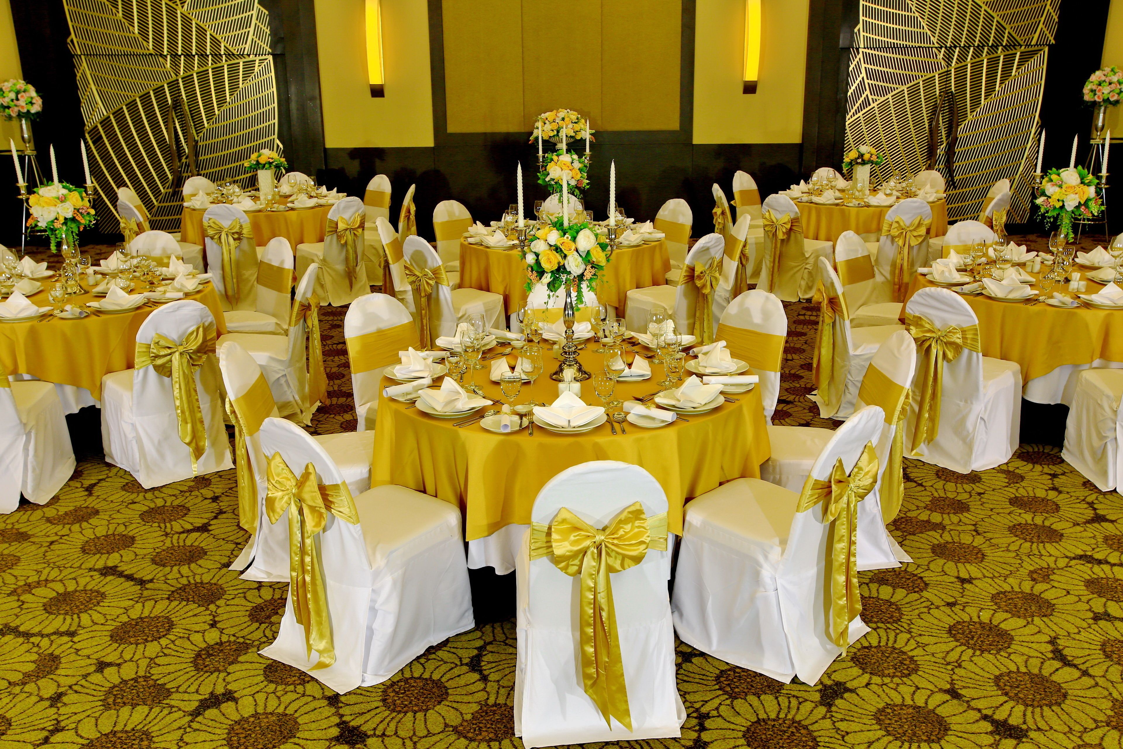 Uma sala de banquetes com mesas redondas dispostas para um evento formal, com toalhas de mesa douradas e brancas, cadeiras com fitas douradas e arranjos florais no centro.