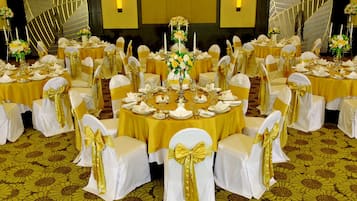 Uma sala de banquetes com mesas redondas dispostas para um evento formal, com toalhas de mesa douradas e brancas, cadeiras com fitas douradas e arranjos florais no centro.