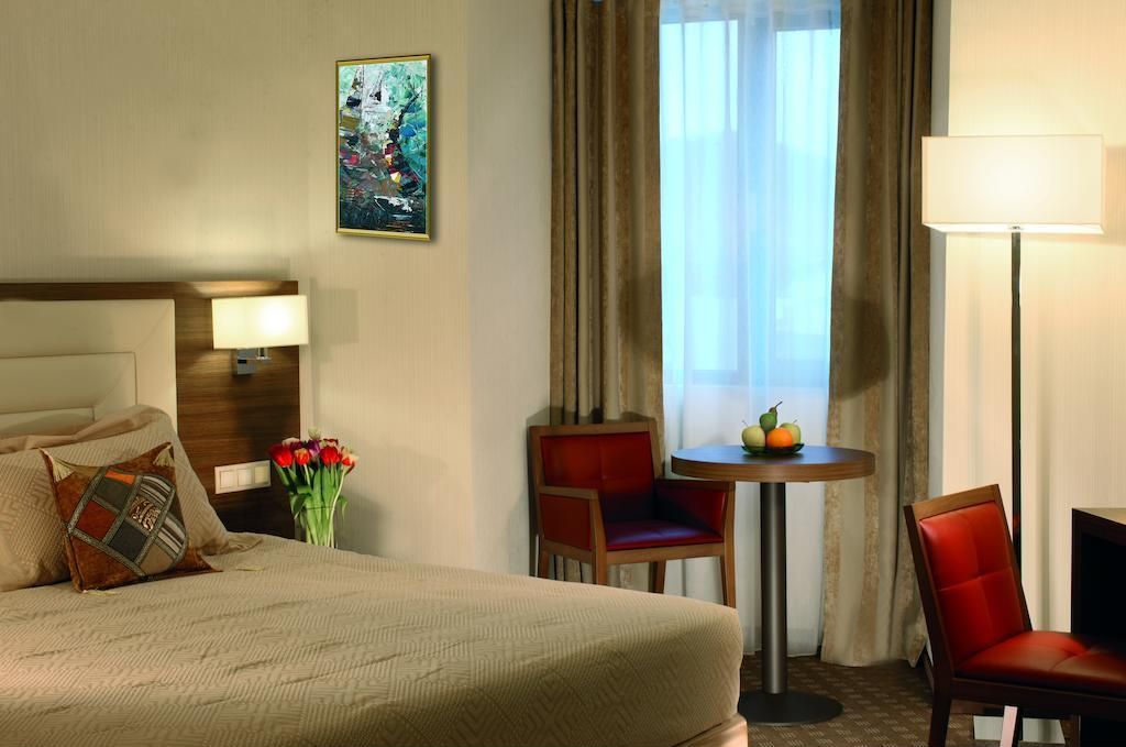 Quarto de hotel com cama, escrivaninha, cadeira, abajur, quadro na parede e janela com cortinas.