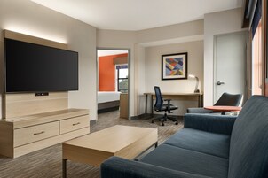 Quarto de hotel moderno com televisão de tela plana, cómoda de madeira, sofá azul, secretária com cadeira e um quadro na parede.