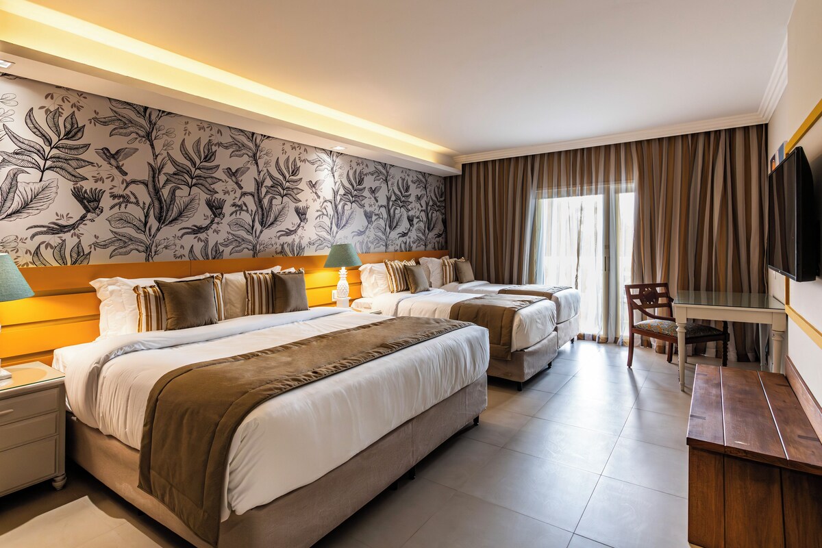 Quarto de hotel com duas camas, uma escrivaninha, uma cadeira, uma televisão e uma parede com padrão floral.