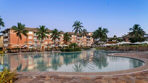 Um hotel com piscina, rodeado por espreguiçadeiras e guarda-sóis, palmeiras e um edifício com várias andares e varandas.