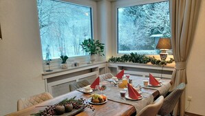Uma área de refeições com uma mesa de madeira posta para quatro pessoas, rodeada por cadeiras. Há detalhes decorativos como uma cesta de frutas e uma pequena planta sobre a mesa.