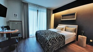 Quarto de hotel com cama, escrivaninha, cadeira, radiador e janela com cortinas.