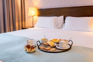 Cama bem arrumada com lençóis brancos, bandeja com itens de café da manhã, incluindo doces, café e frutas, e um abajur emitindo iluminação suave.
