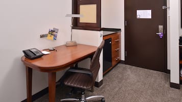 Quarto de hotel com uma mesa de madeira, telefone, abajur, cadeira, armário e porta com fechadura e orifício para chave.