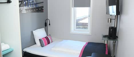 Quarto de hotel compacto com cama, escrivaninha, cadeira, janela com vista, televisão na parede e um cabideiro.
