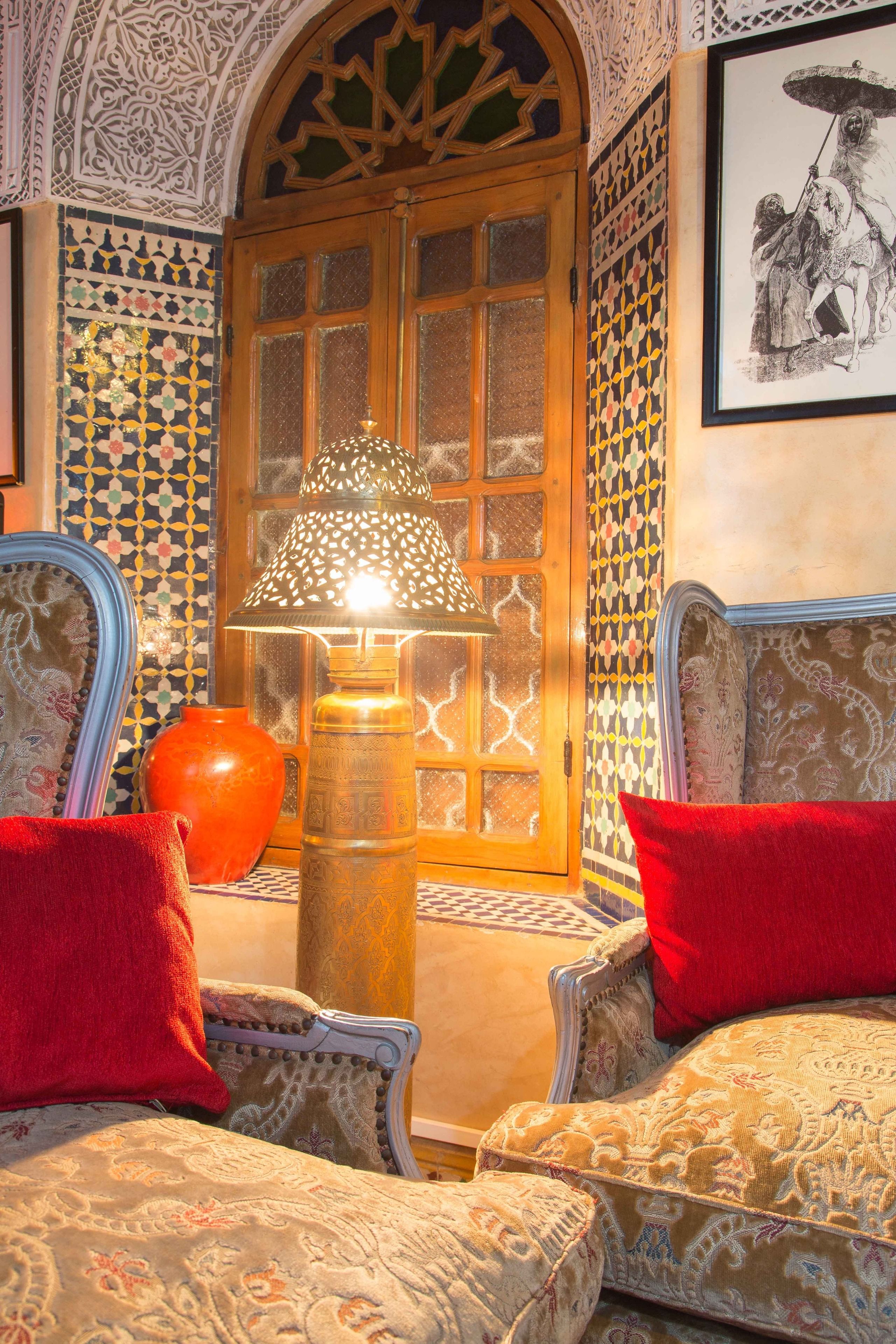 Quarto com paredes em azulejos decorativos, porta de madeira com painéis de vidro, sofá estampado com almofadas vermelhas e uma luminária de mesa.