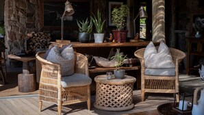 Uma área de estar acolhedora ao ar livre, com cadeiras de vime e uma mesa redonda de madeira, cercada por plantas em vasos e uma parede de pedra.