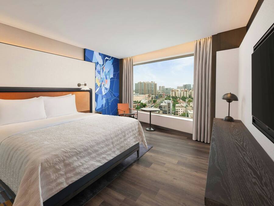 Quarto de hotel moderno com uma cama grande, televisão, uma mesinha e vista para a cidade.
