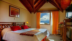 Quarto de hotel com uma cama grande, cabeceira de madeira, um quadro na parede, uma janela com cortinas e uma TV em um móvel de madeira.