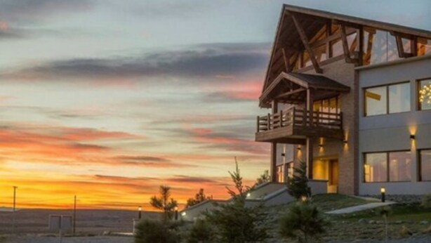 Um edifício moderno de dois andares com varanda no andar superior, localizado numa encosta com vista para o oceano ao pôr do sol.