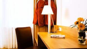 Quarto de hotel com escrivaninha de madeira, uma cadeira, um abajur, um casaco pendurado em um suporte e um vaso com flores.