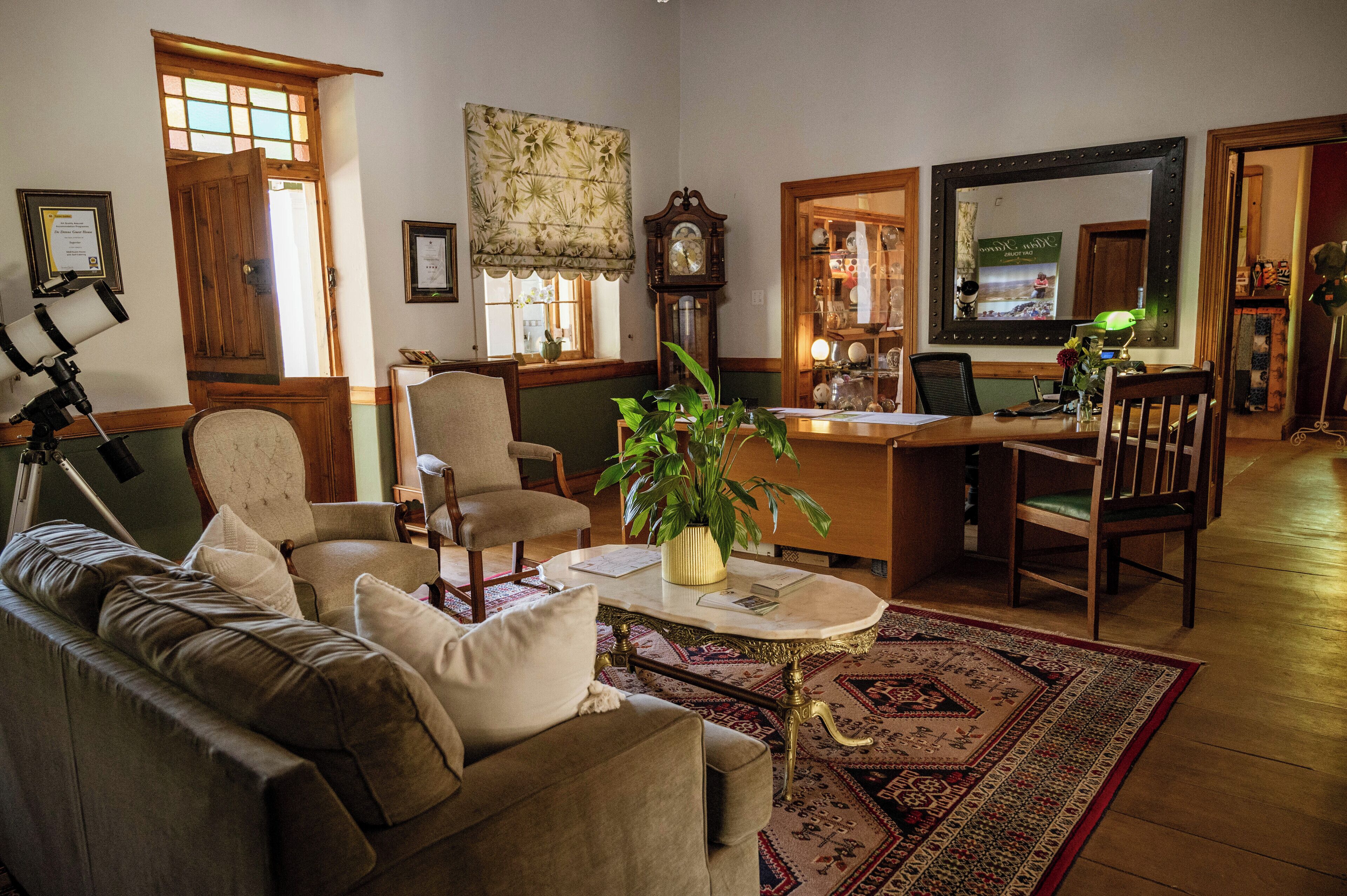 Uma sala de estar com sofá, cadeiras, mesa de centro, planta, relógio e uma área de jantar com mesa e cadeiras.