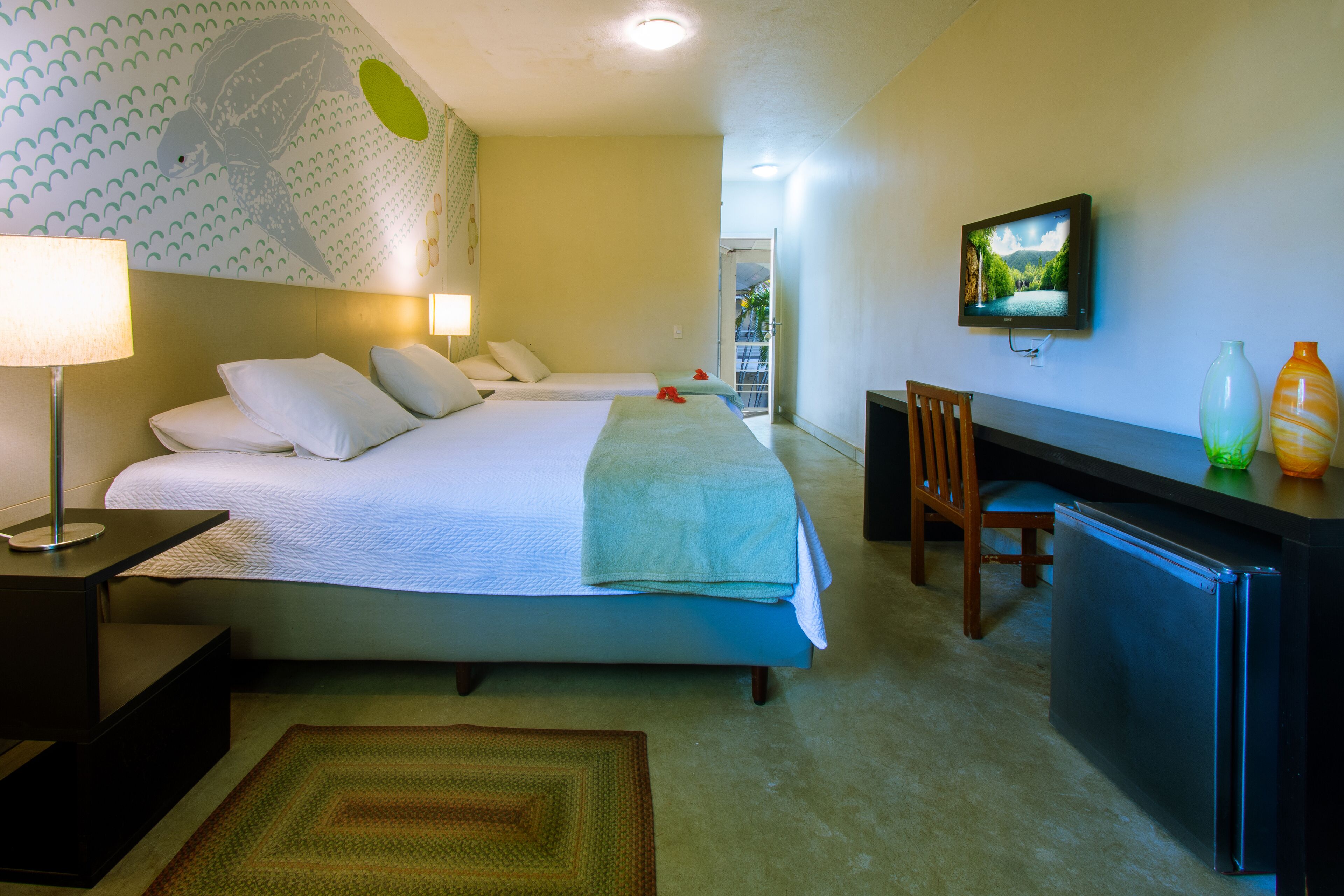 Quarto de hotel com duas camas, uma escrivaninha, uma cadeira, uma televisão e vista para o exterior.