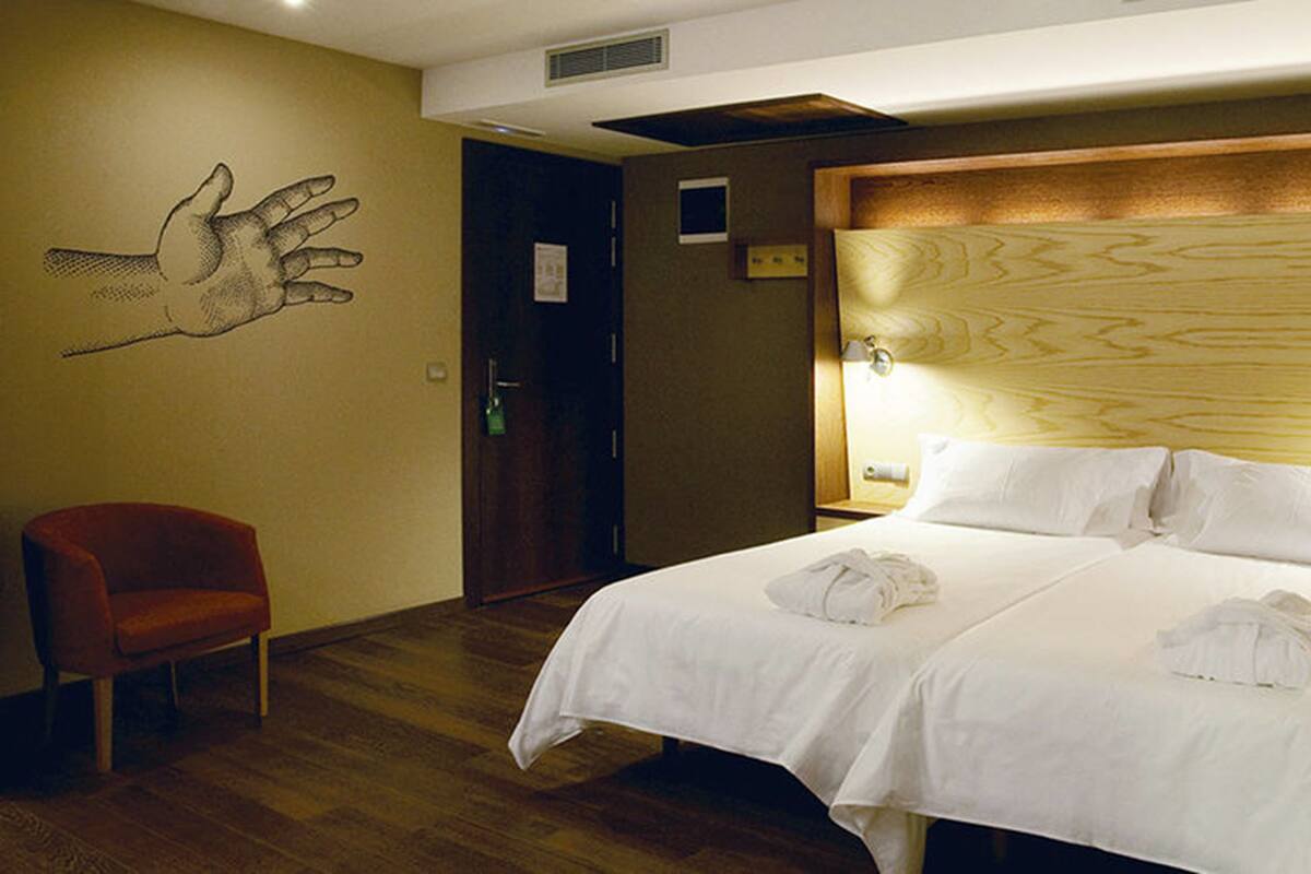Quarto de hotel com uma cama grande, duas mesas de cabeceira com luminárias, uma cabeceira de madeira, uma cadeira vermelha e uma obra de arte na parede.