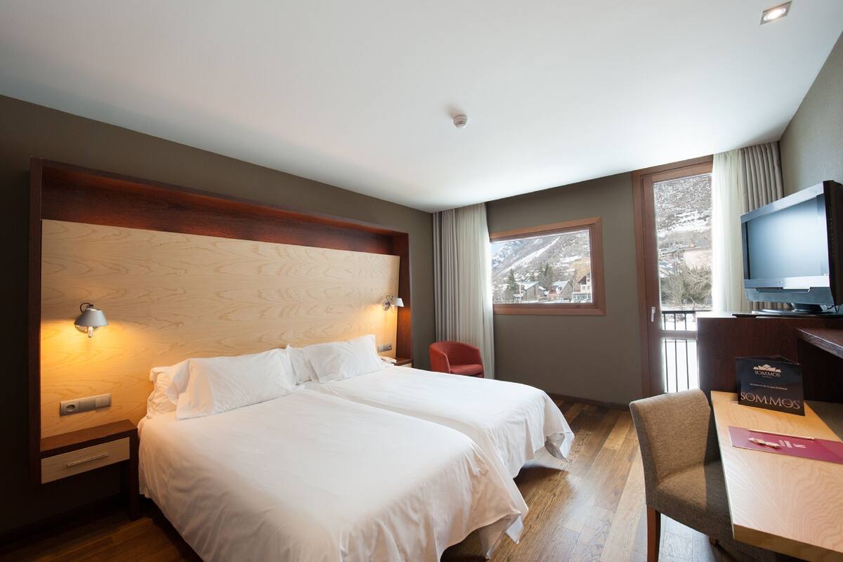 Quarto de hotel com duas camas, uma escrivaninha, televisão e janela com vista para árvores.