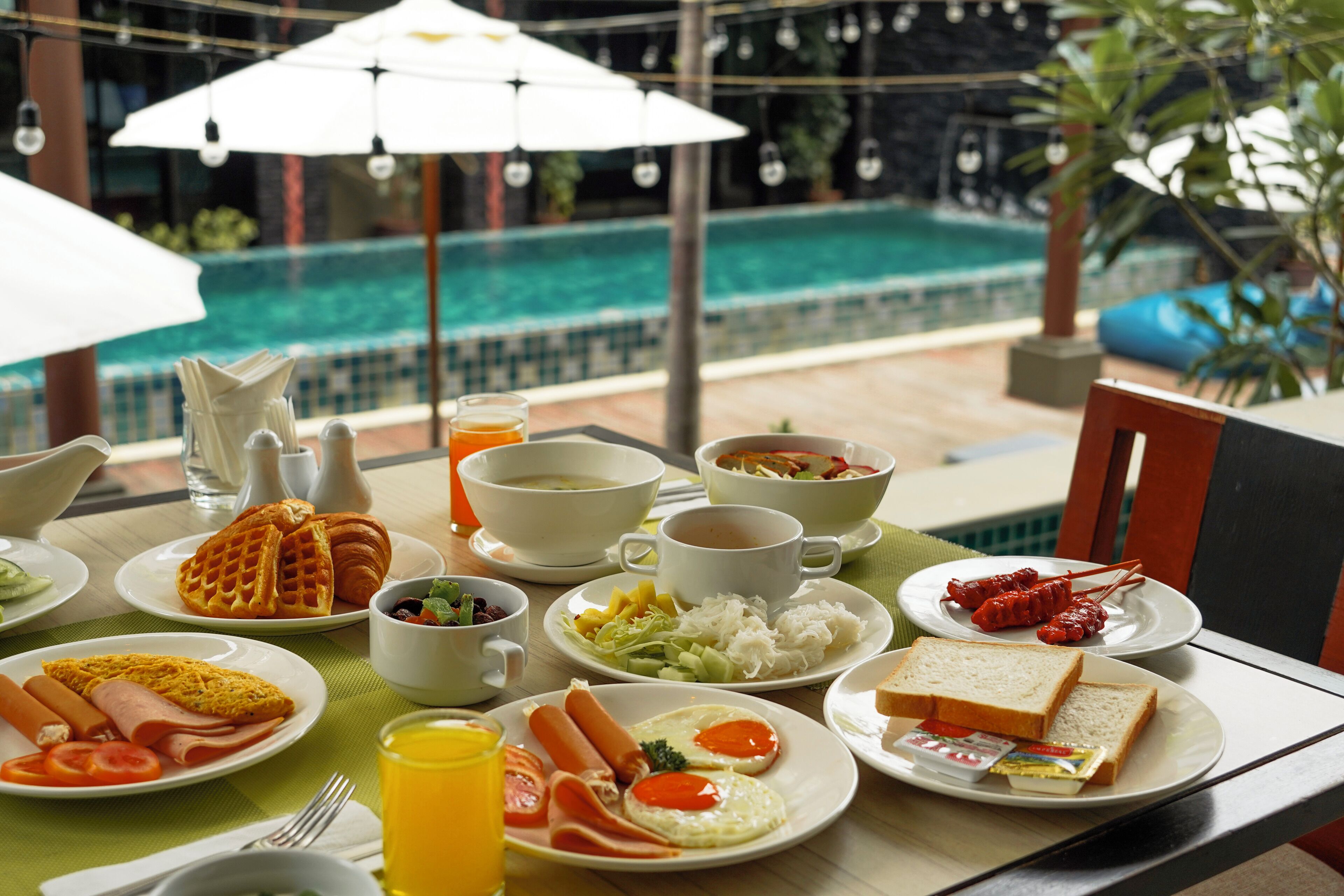Buffet de café da manhã com diversos pratos, piscina ao fundo e guarda-sóis para proteger do sol.