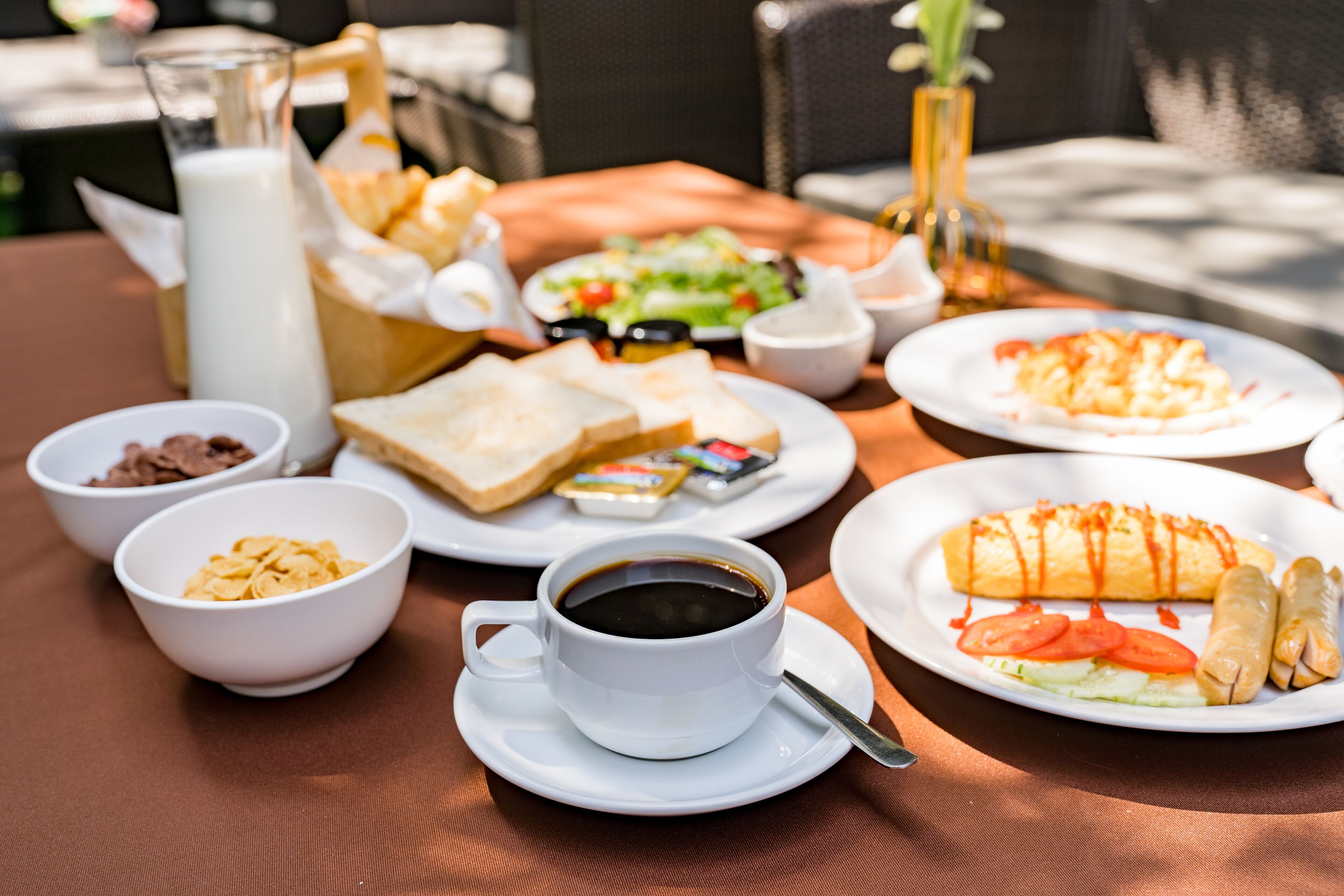 Uma mesa com diversos itens de pequeno-almoço, incluindo torradas, uma chávena de café, uma salada e um prato com omelete recheada e legumes.