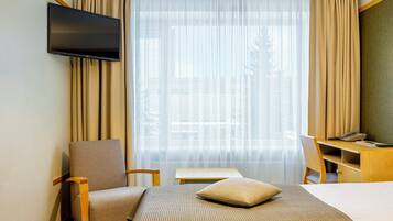 Quarto de hotel com cama, escrivaninha, cadeira, televisão e janela com cortinas.