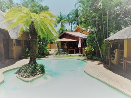 Piscina rodeada por vegetação tropical e um edifício de madeira com telhado de palha.