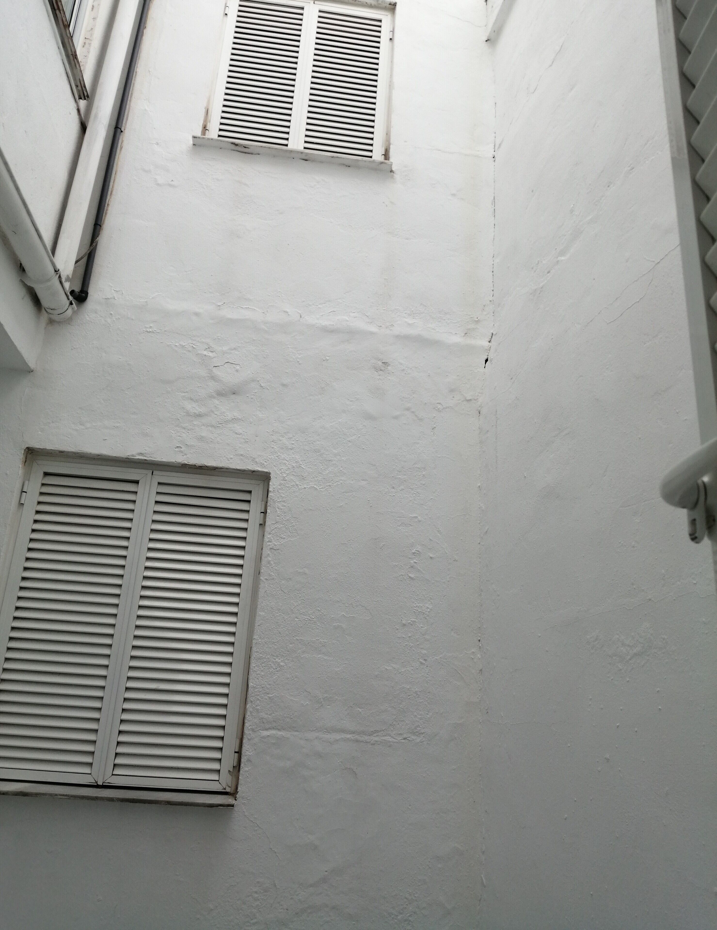 Fachada de edifício com duas janelas brancas com persianas, um cano visível e uma parede texturizada.